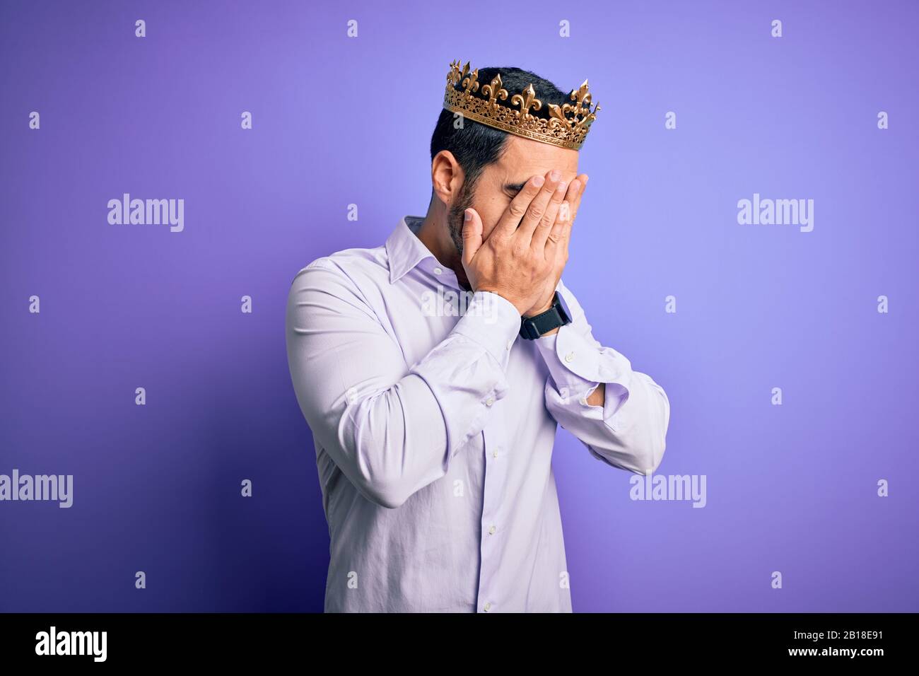 Young handsome man with beard wearing golden crown of king over purple ...