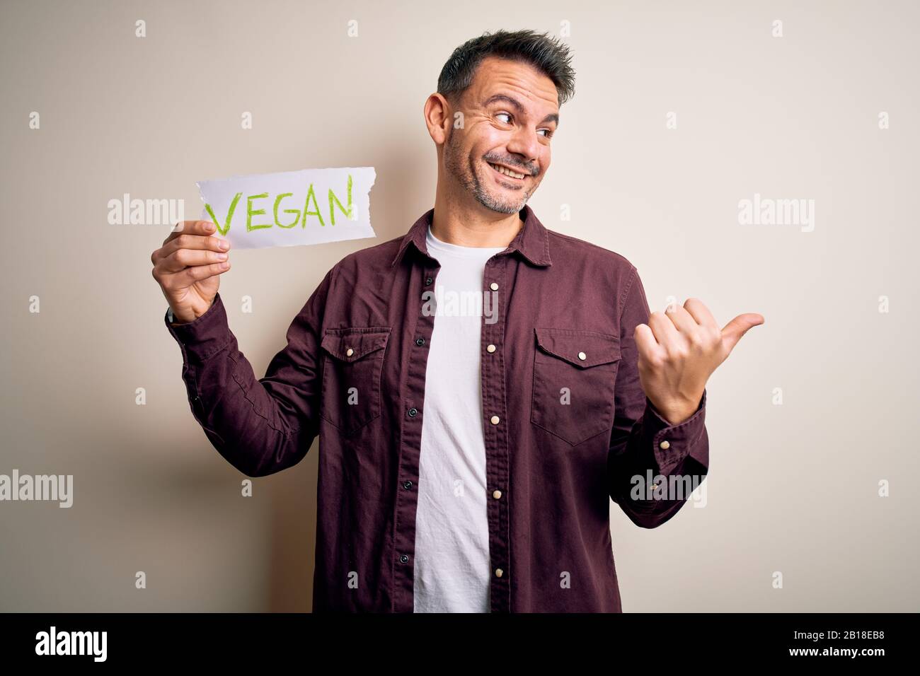 Young handsome veggie man holding paper with vegan message over white ...