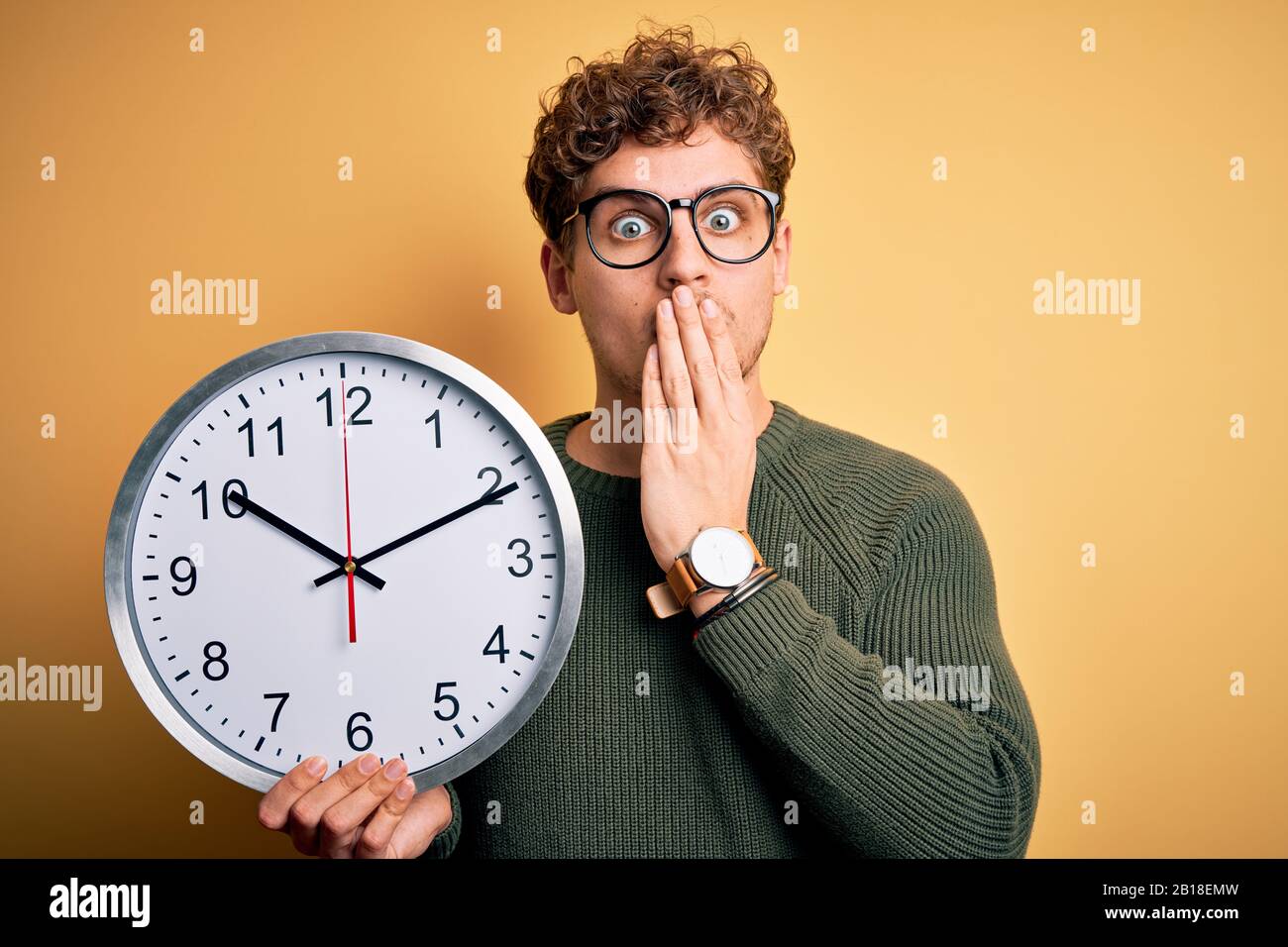 Young blond man with curly hair wearing glasses holding big clock over ...