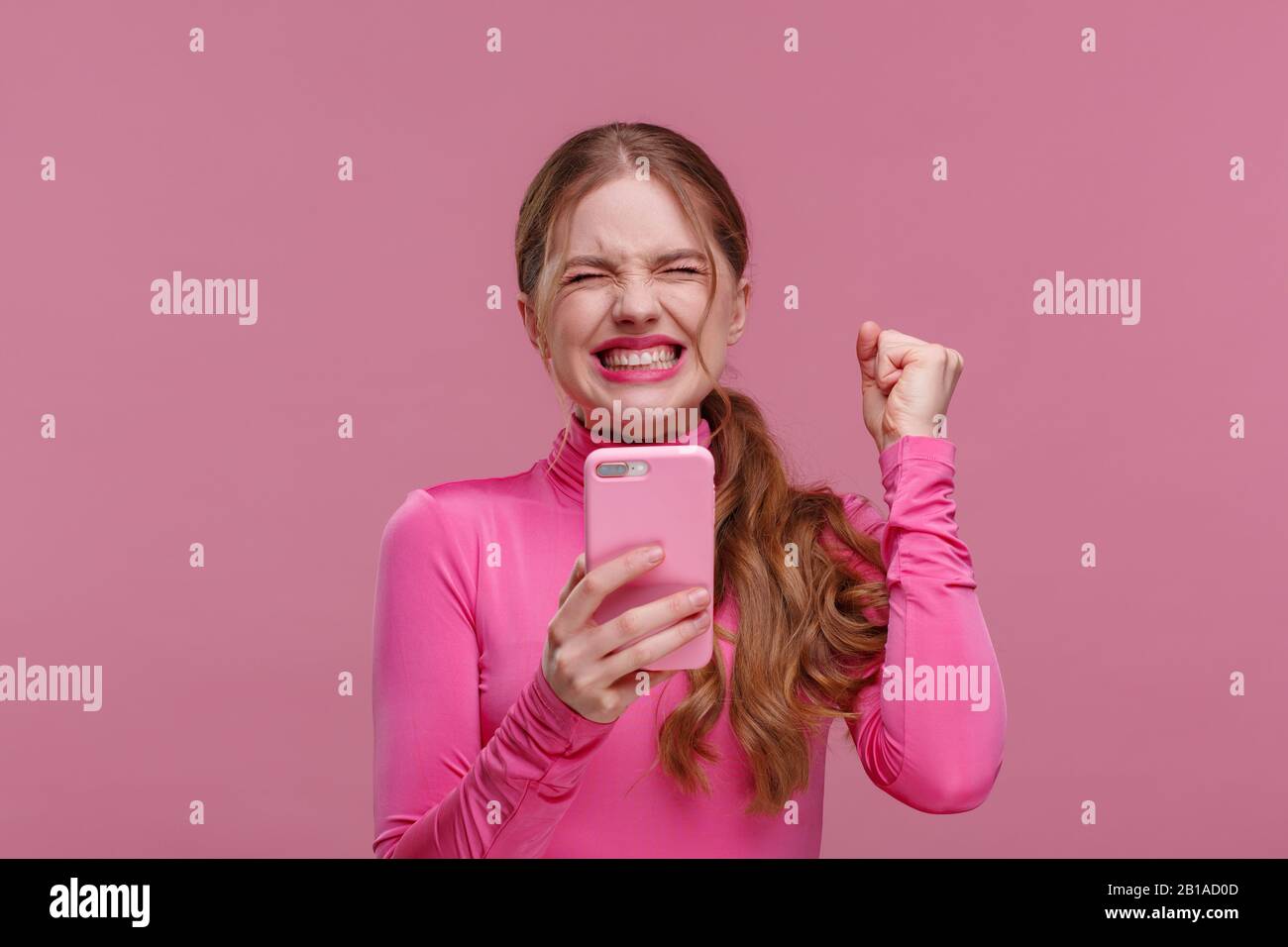 The best day ever. Surpised young redhead woman holding pink smartphone ...