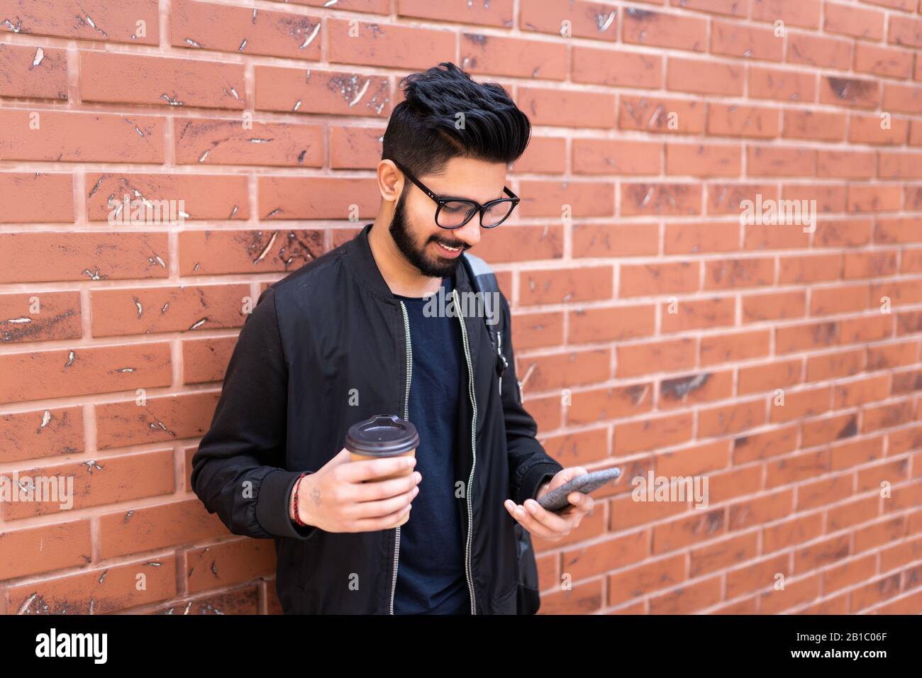 Handsome indian man use mobile phone enjoying coffee against brick wall ...