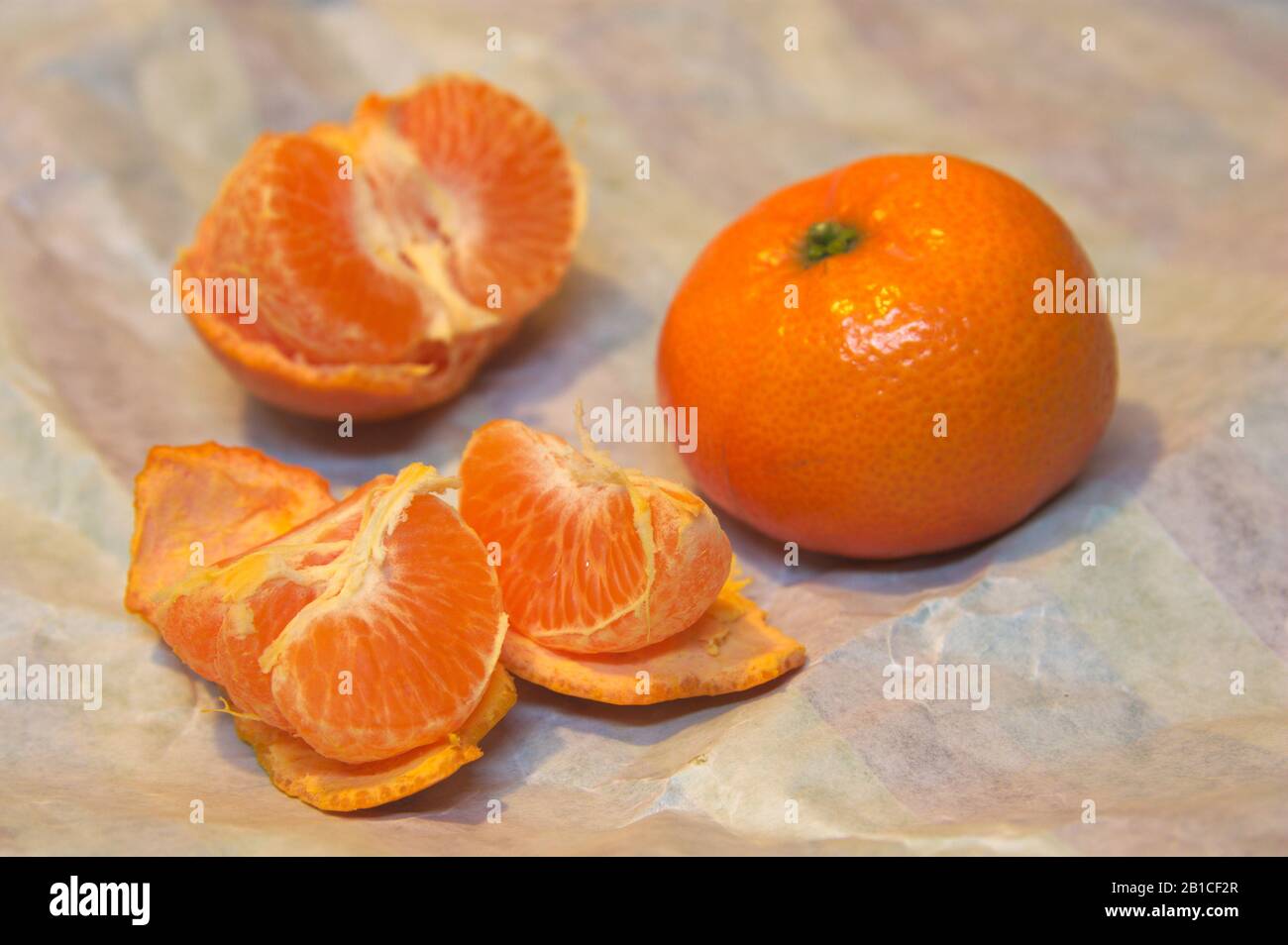A tangerine peeled and open next to another with skin on a crumpled ...