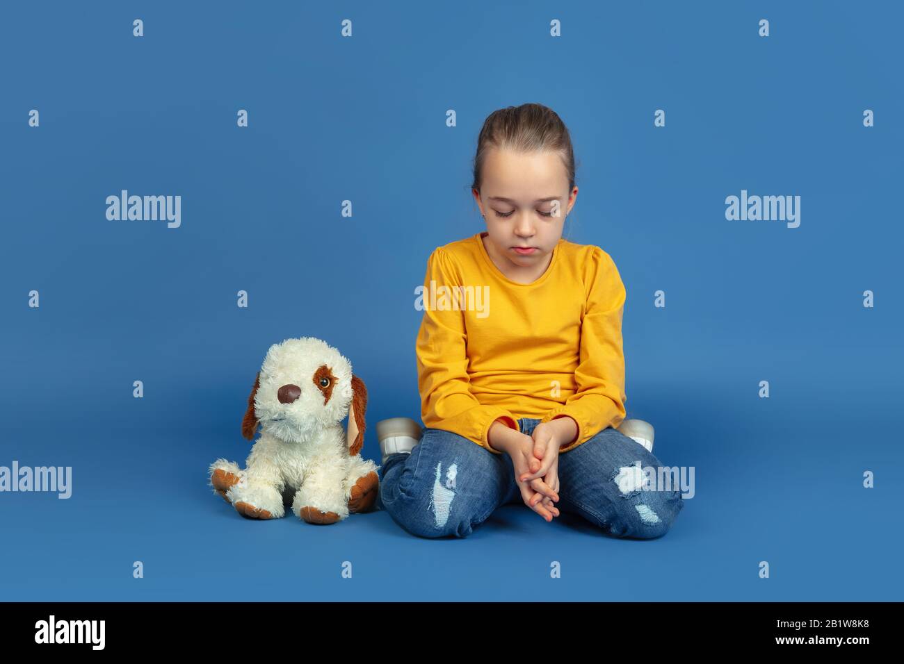 Portrait of sad little girl sitting isolated on blue studio background ...