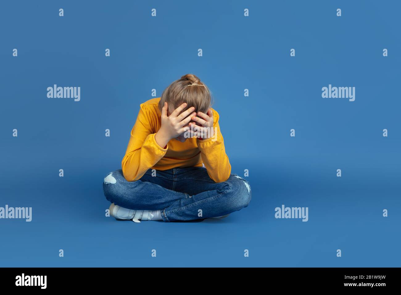 Portrait of sad little girl sitting isolated on blue studio background ...
