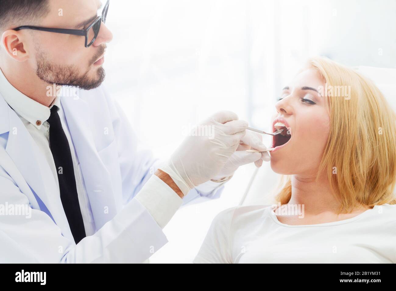 Young handsome dentist examining teeth of happy woman patient sitting on dentist chair in dental ...