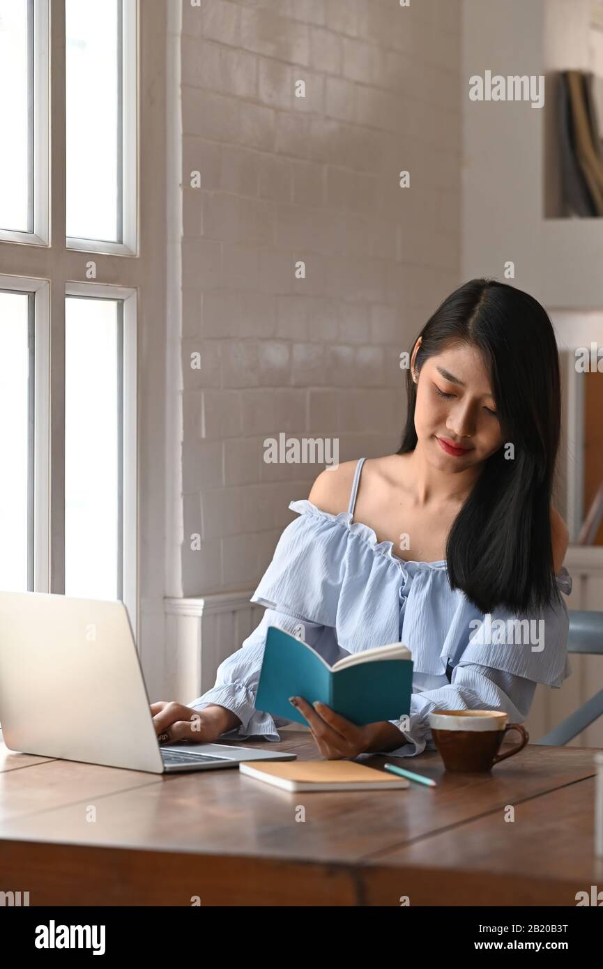 Beautiful woman reading a book while sitting in front a computer laptop ...