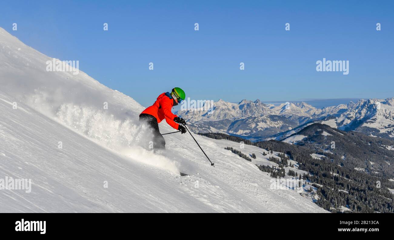 Skier going down steep slope, black piste, blue sky, mountains behind ...