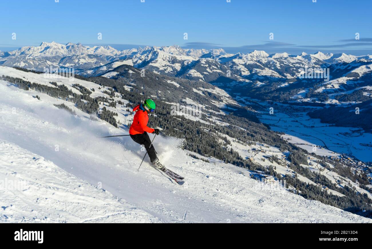 Skier going down steep slope, black piste, blue sky, mountains behind ...