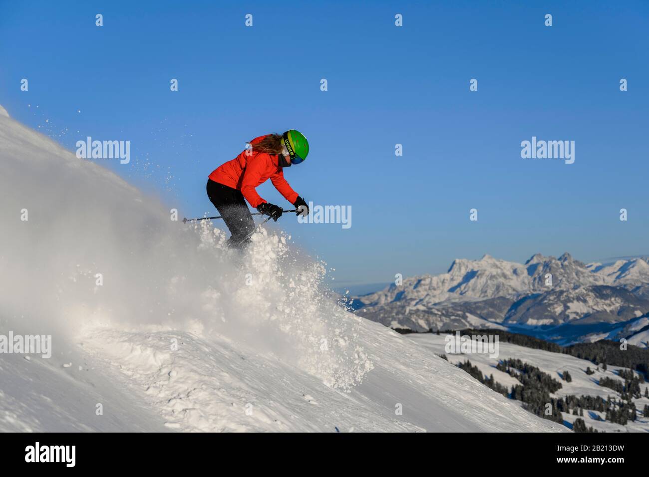 Skier going down steep slope, black piste, blue sky, mountains behind ...