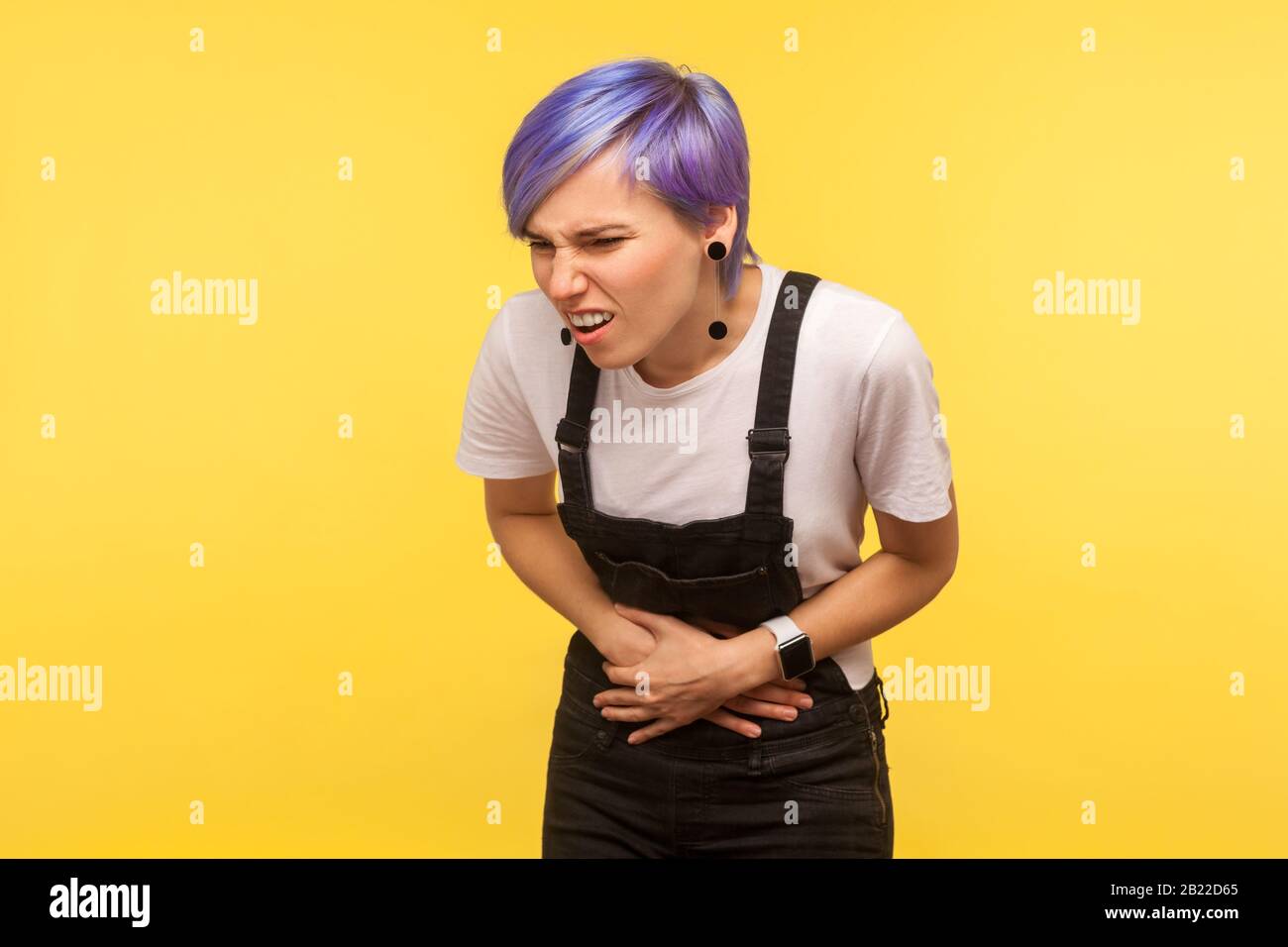 Stomachache. Portrait of ill woman with violet short hair in overalls ...