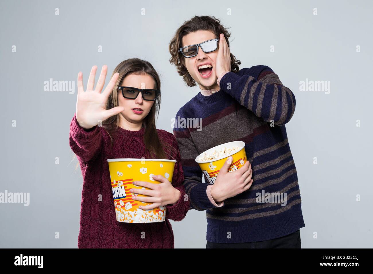 Young scared couple, woman and man in 3d glasses watching movie film on date holding buckets of ...