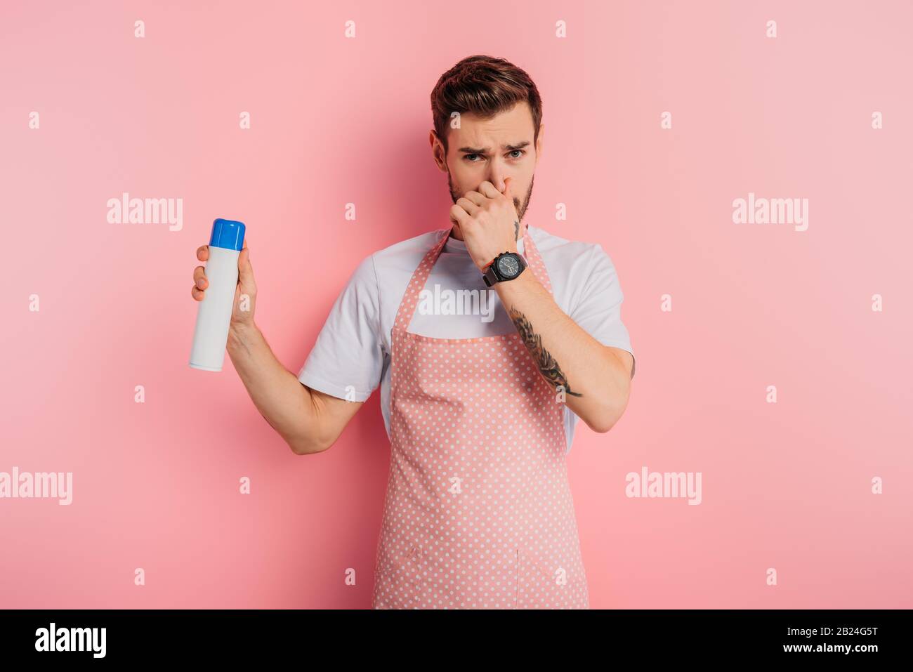 displeased young man in apron plugging nose with hand while holding air ...
