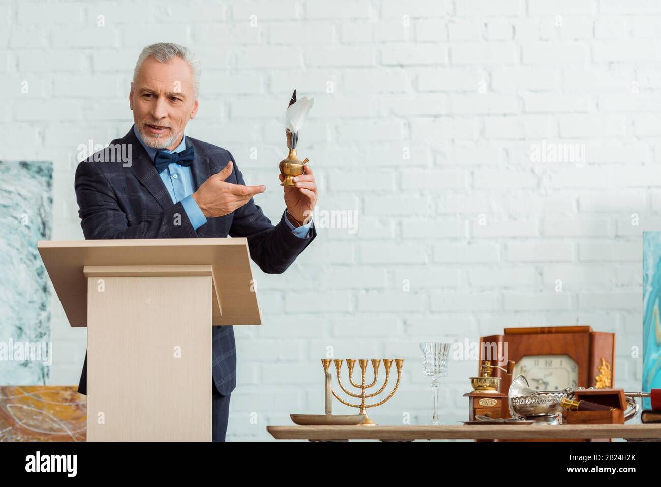 handsome auctioneer in suit pointing with hand at feather pen during ...