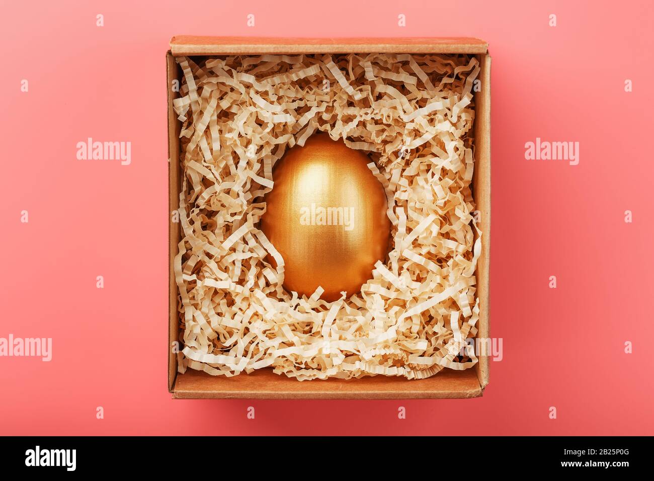 Golden egg in a cardboard box with shavings on a pink background. The ...
