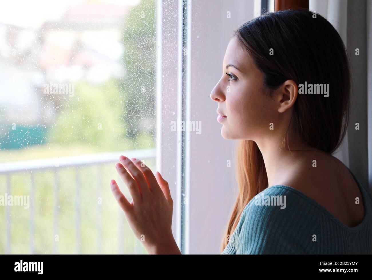 Melancholic sad young woman looking through the window at home in a ...