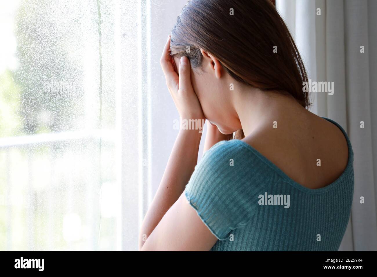 Sad depressed girl crying near the window at home in a rainy day, focus ...