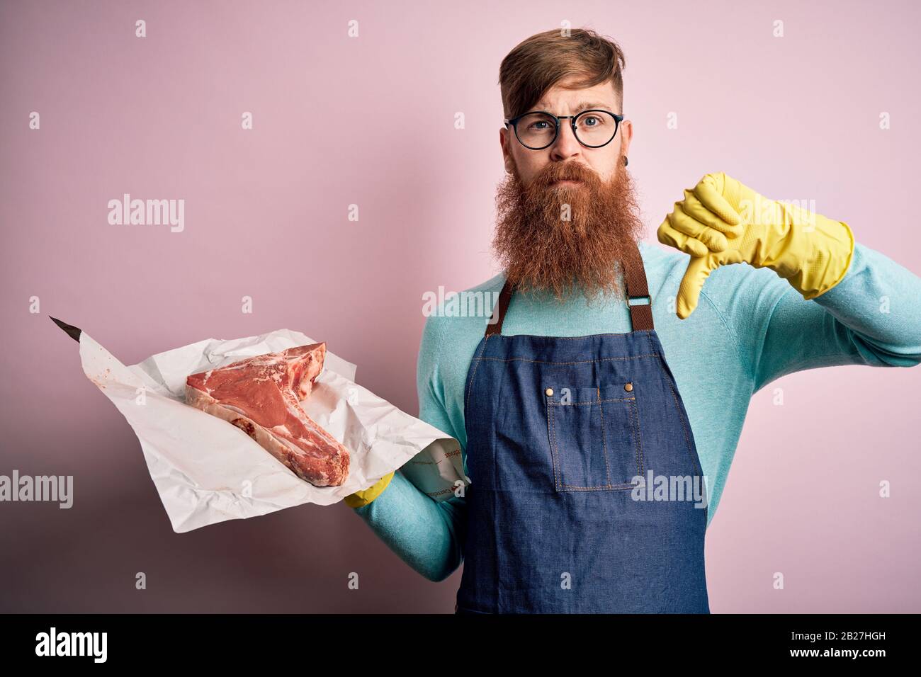 Redhead Irish butcher man with beard holding raw beef steak over pink ...