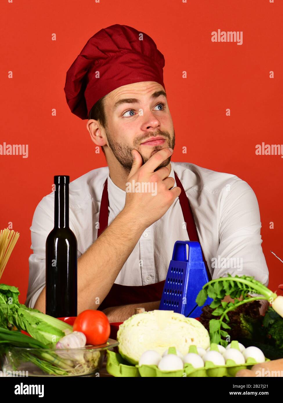 Chef with curious face sits by food on red background. Vegetarian meal ...