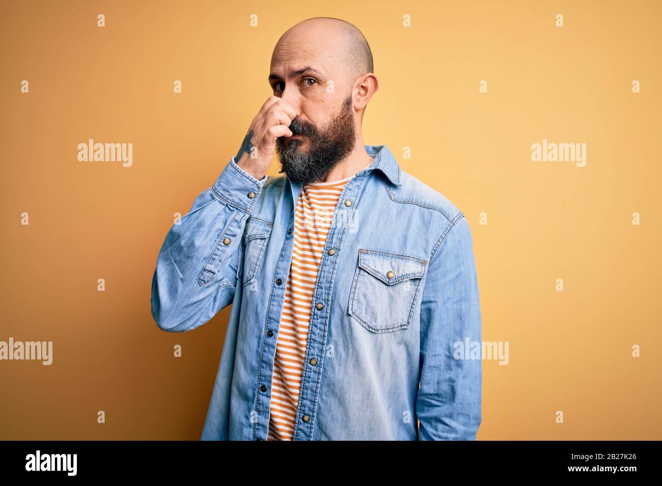 Handsome bald man with beard wearing casual denim jacket and striped t-shirt smelling something ...