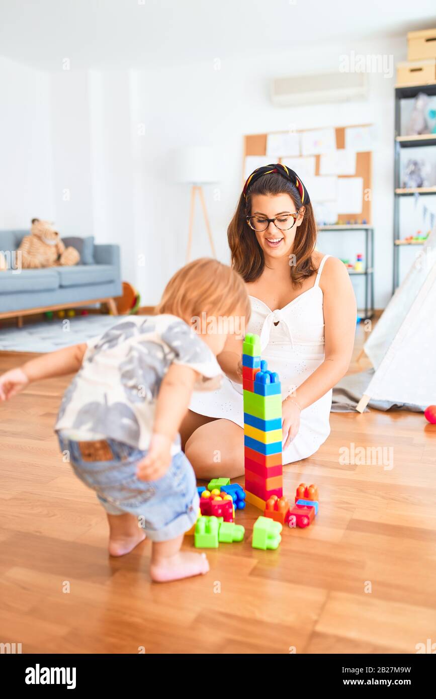 Beautiful teacher and toddler playing with building blocks around lots of toys at kindergarten ...