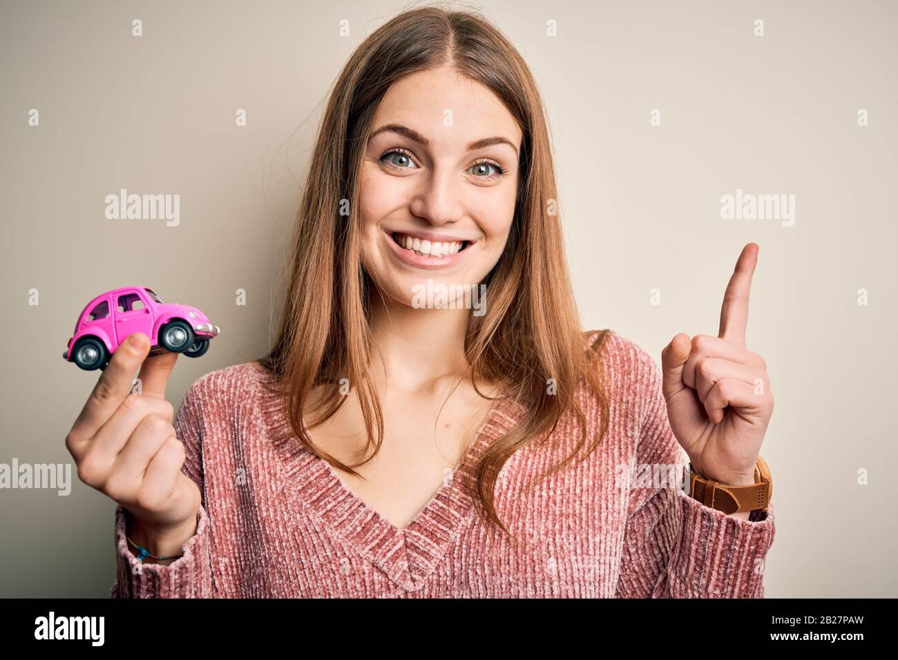 Young beautiful redhead woman holding small pink car over isolated ...