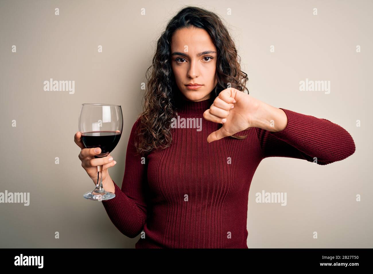 Young beautiful woman with curly hair drinking glass of red wine over ...
