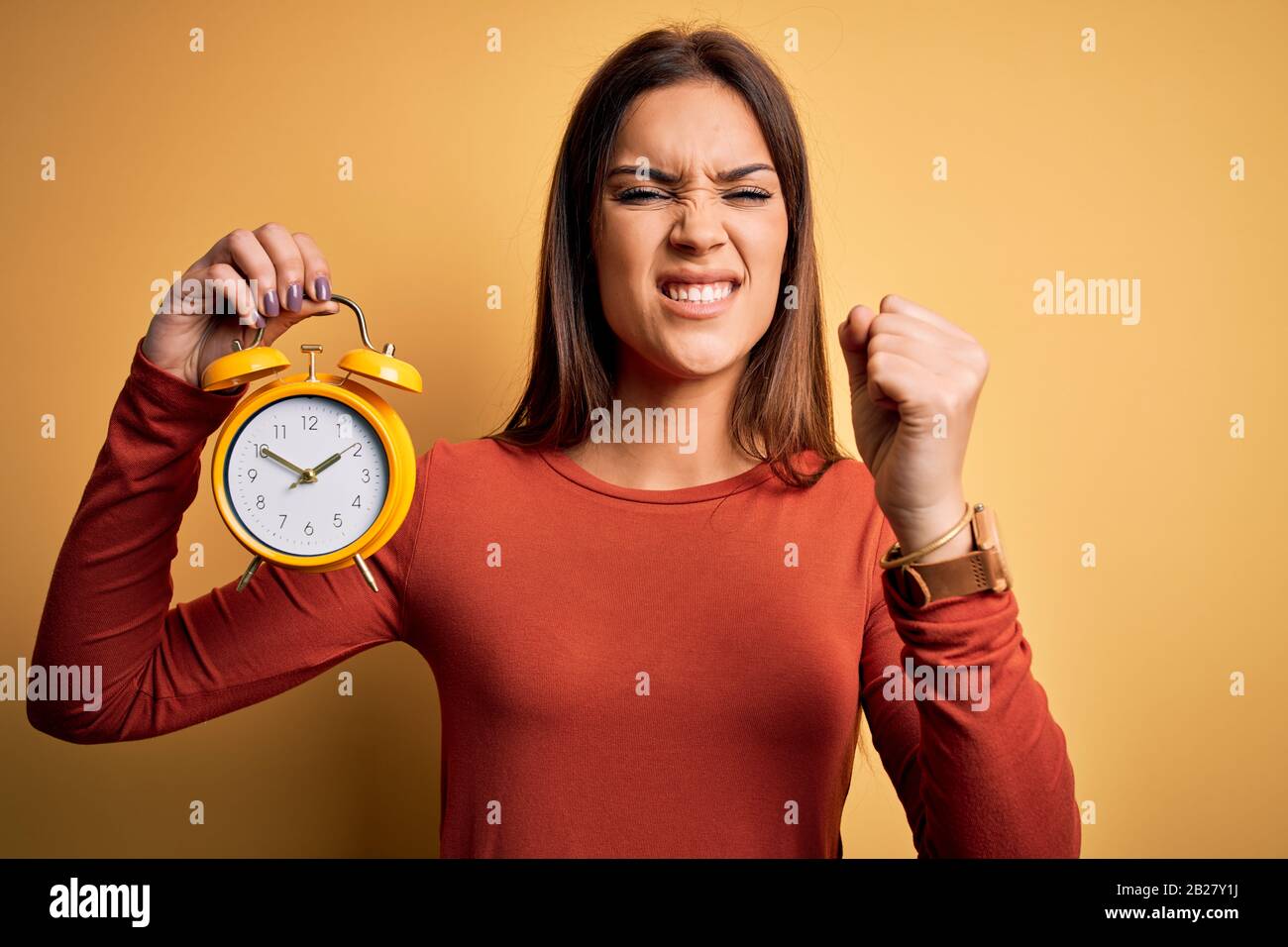 Young beautiful brunette woman holding alarm clock over isolated yellow ...