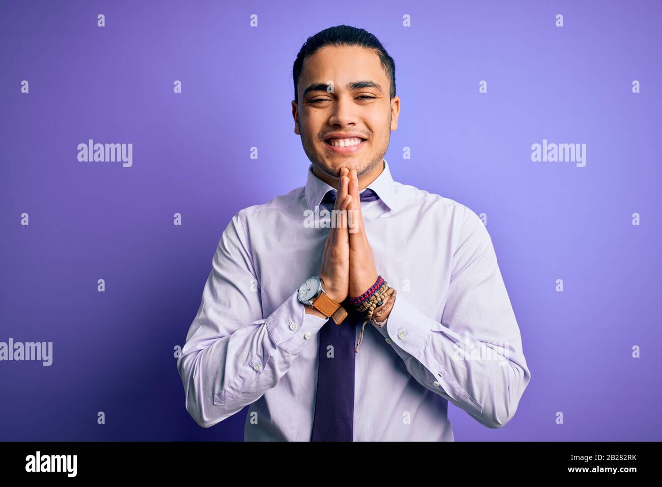 Young brazilian businessman wearing elegant tie standing over isolated purple background praying ...