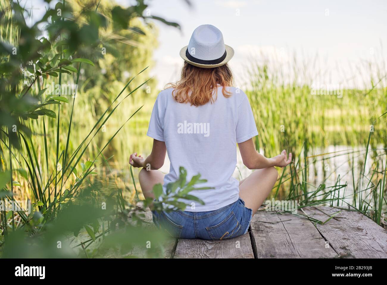 Young woman sitting in lotus position on bridge near water, back view ...