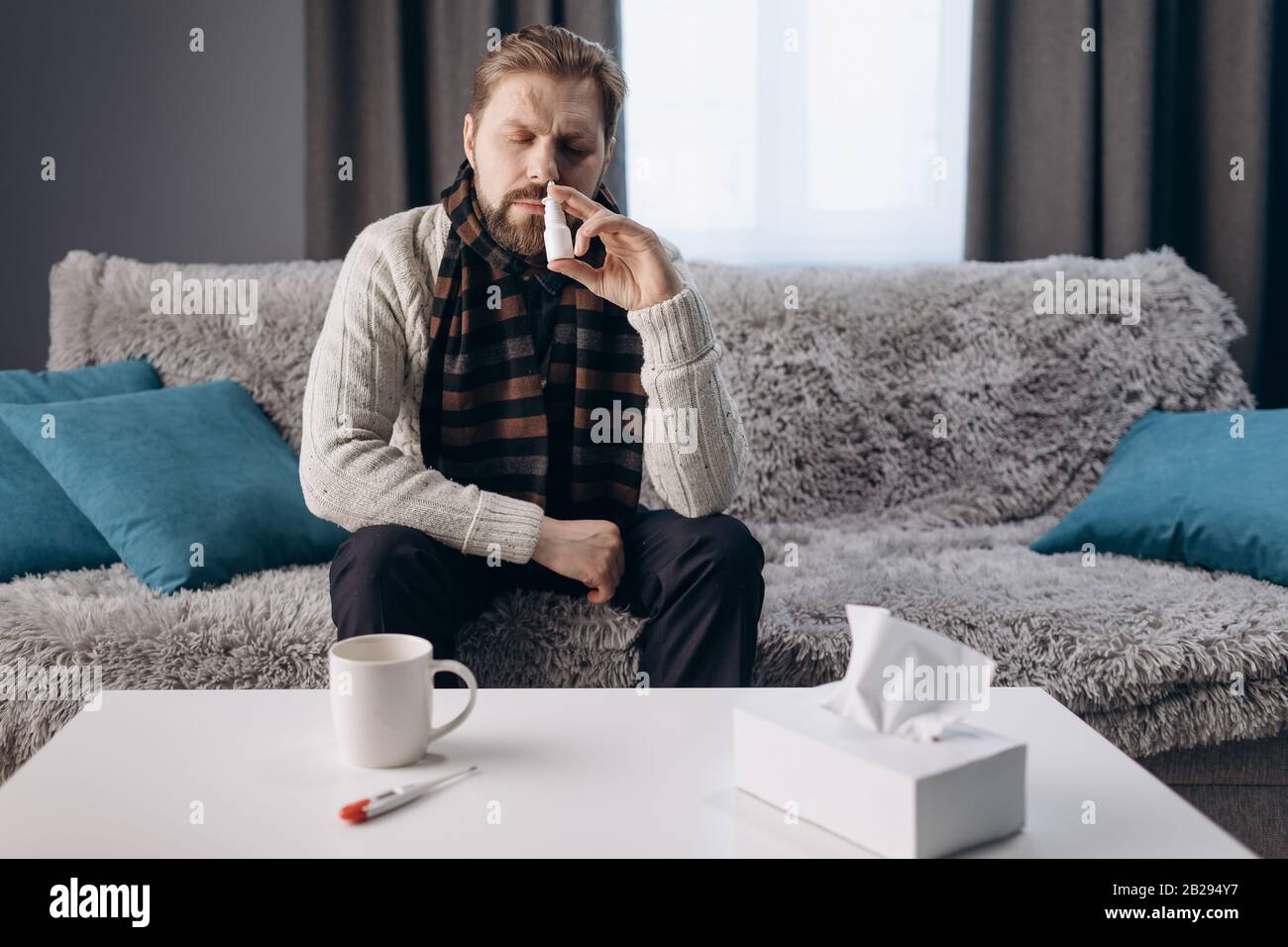 Mature sick man in warm home clothing and knitted scarf sitting on sofa ...