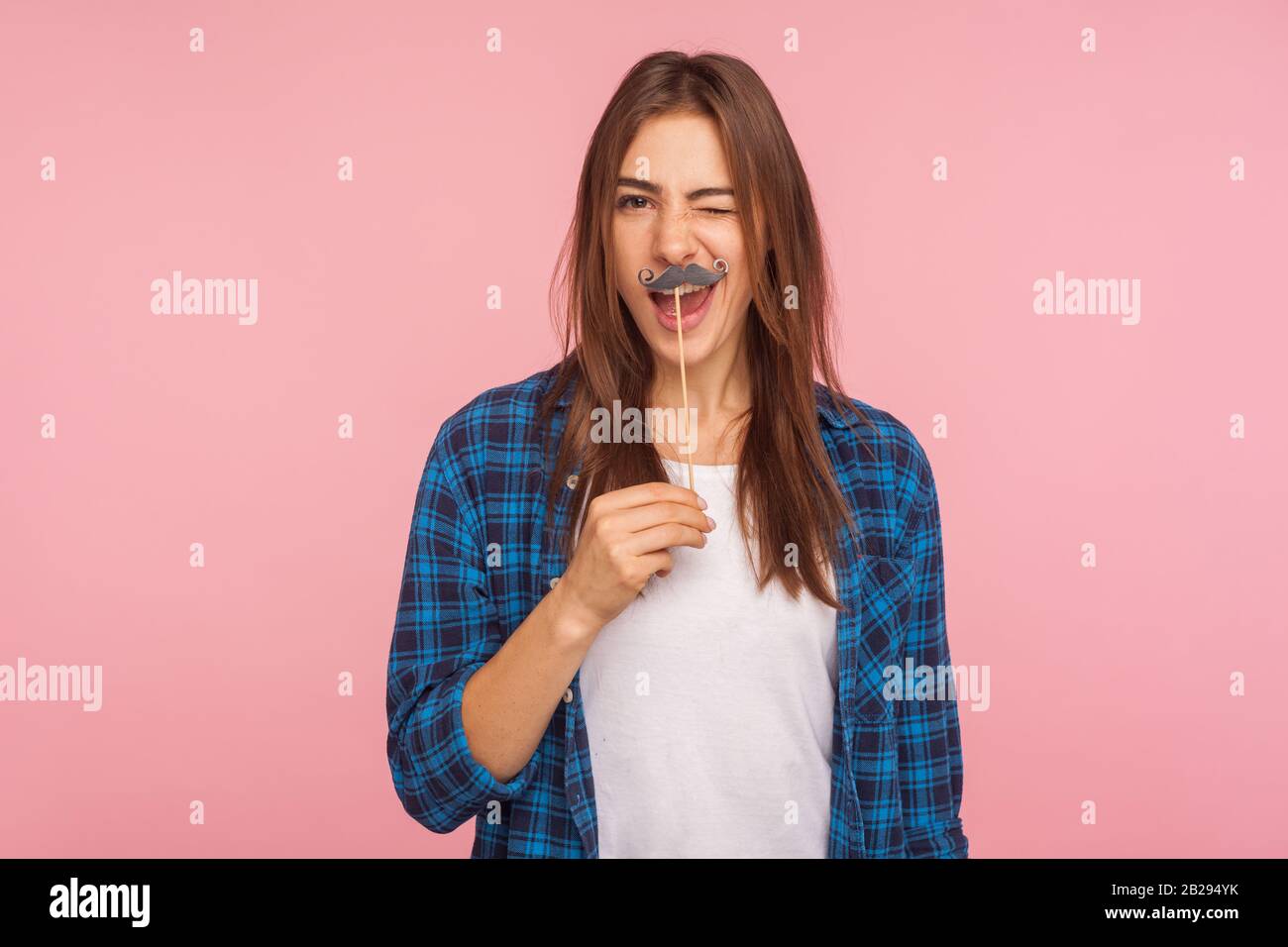 Portrait of positive playful girl in checkered shirt holding fake curled mustache on stick and ...