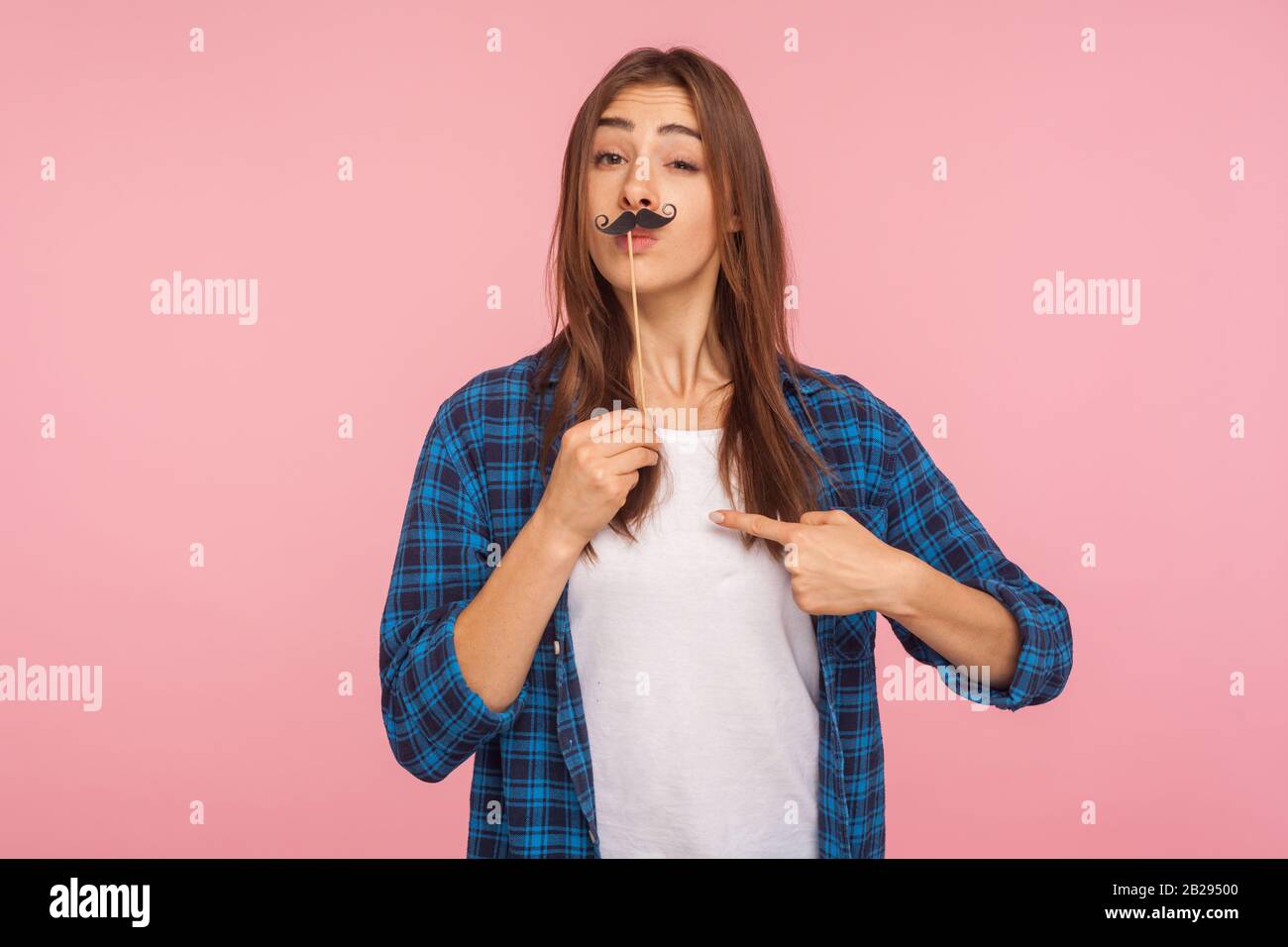 Portrait of playful girl in checkered shirt holding fake curly mustache on stick and pointing ...