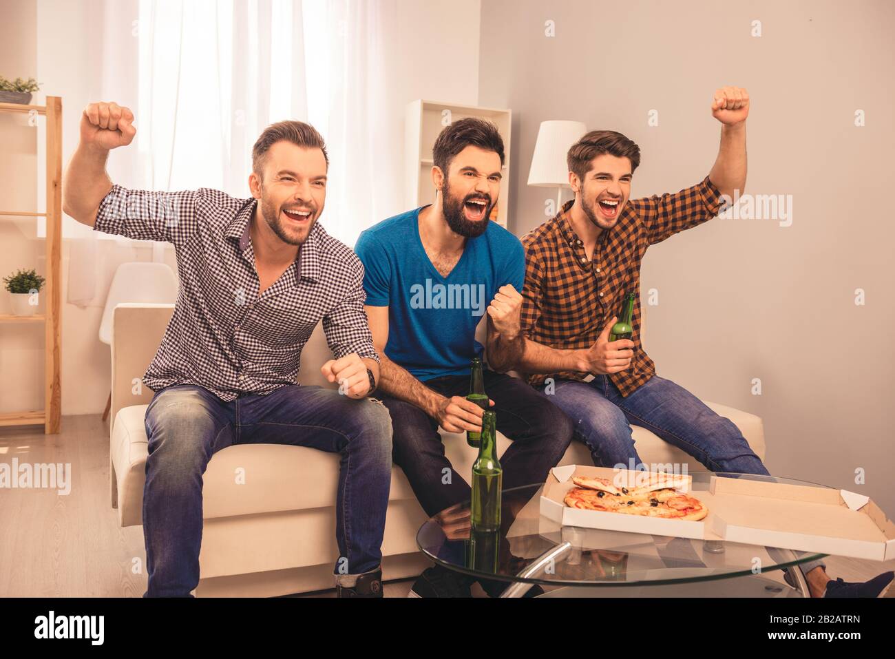 Portrait of three happy men holding bottle of beer and watching tv ...