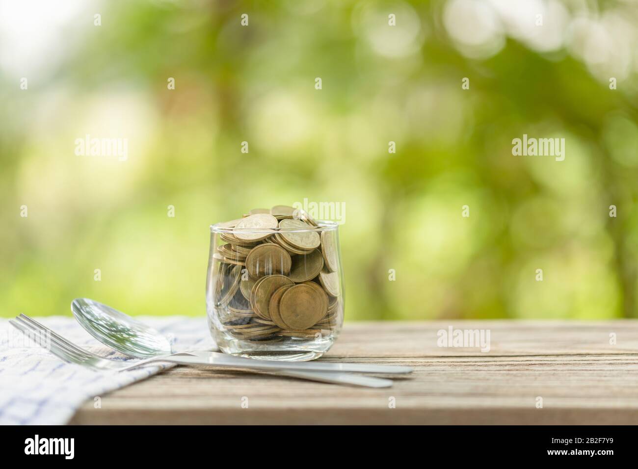 Close up coins in clear money jar, fork and spoon on wooden table with ...