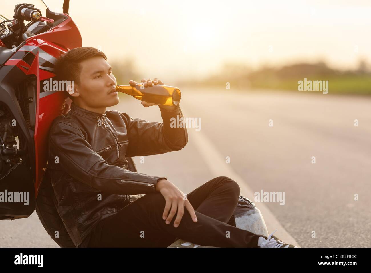 Motorcyclist sitting on the road beside his motorcycle and drinking an ...