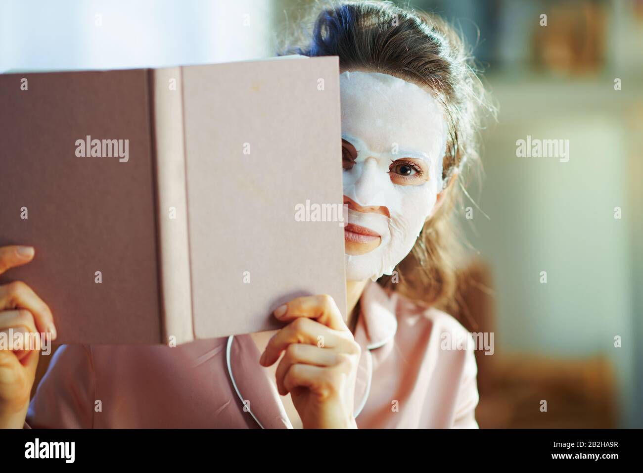elegant female in pajamas with white sheet facial mask looking out from ...