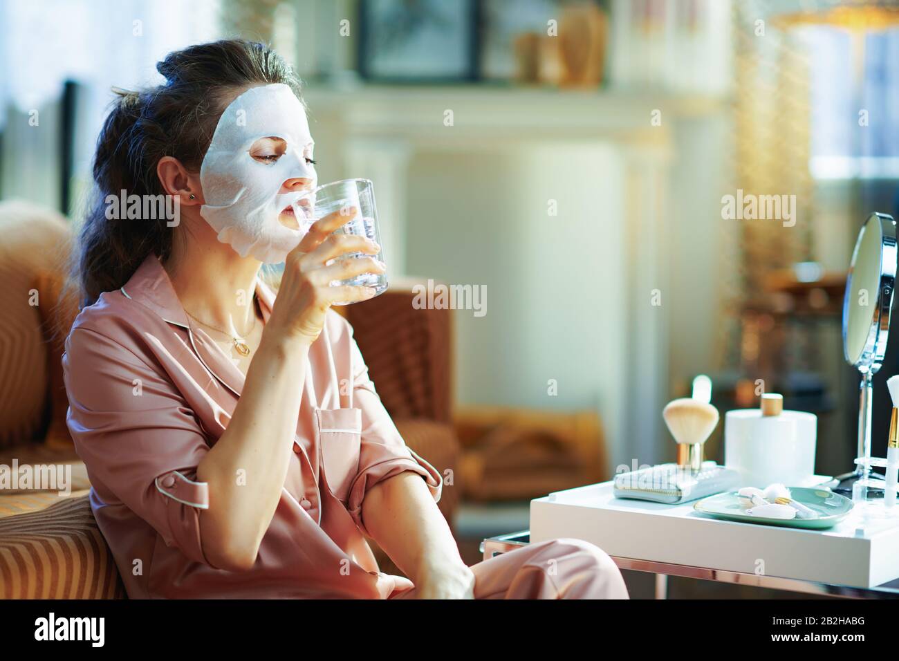 elegant woman in pajamas with white sheet facial mask on face drinking ...