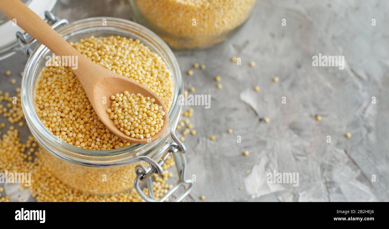 Raw dry hulled millet in a glass jar with a spoon on a grey background ...