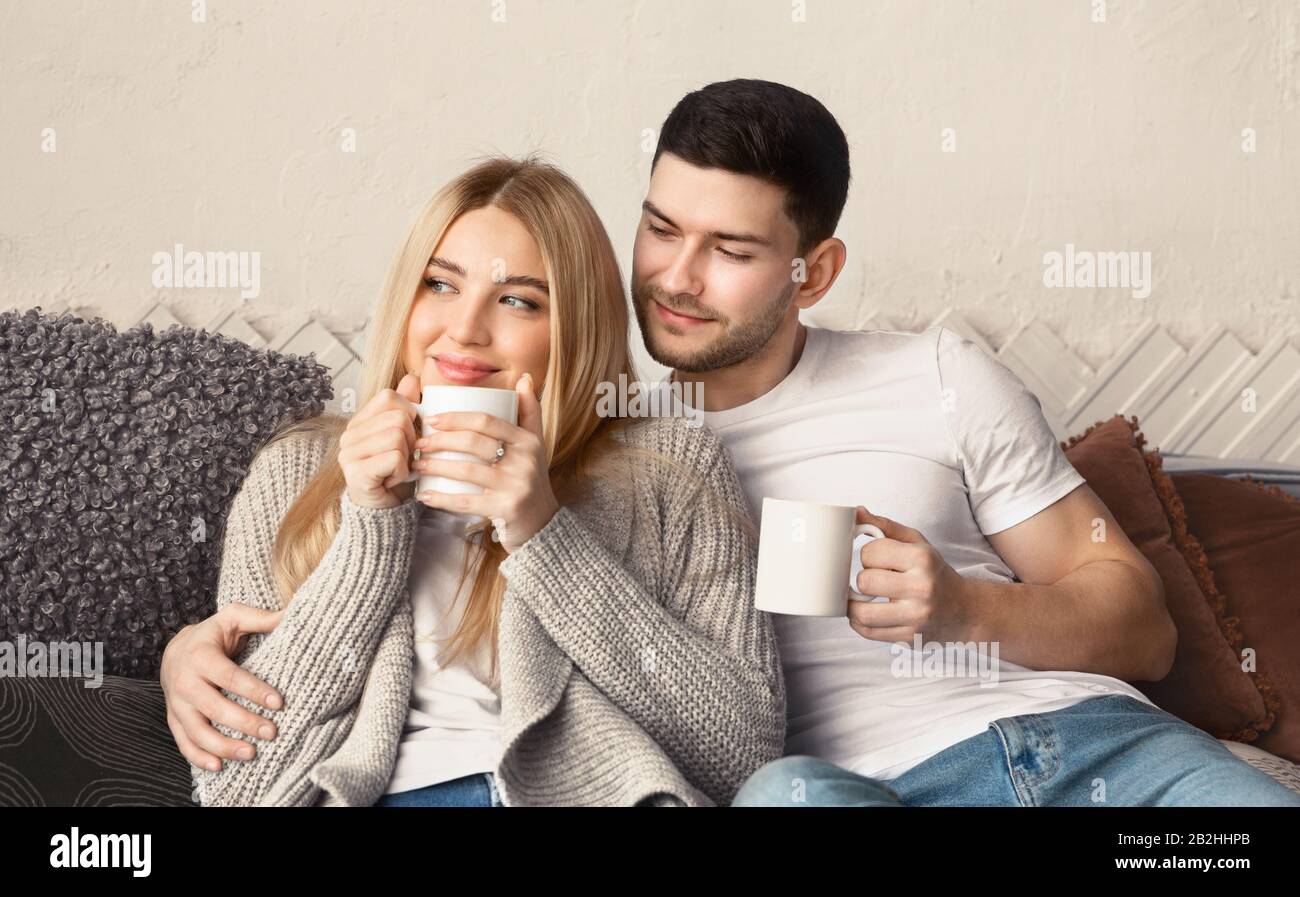Affectionate couple with hot drinks cuddling on sofa in living room ...