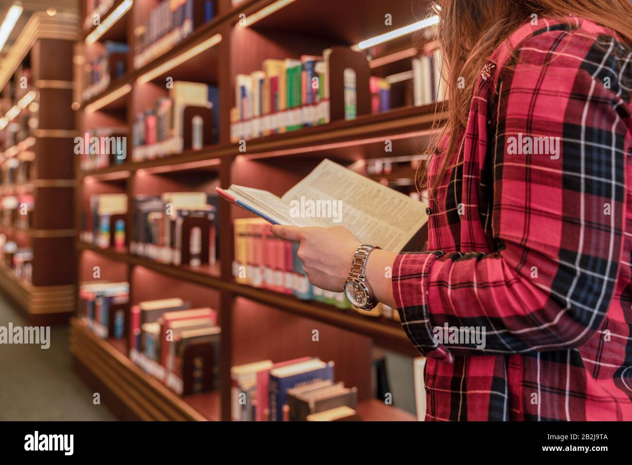 Rear view of student girl standing in library in front of the bookshelf ...
