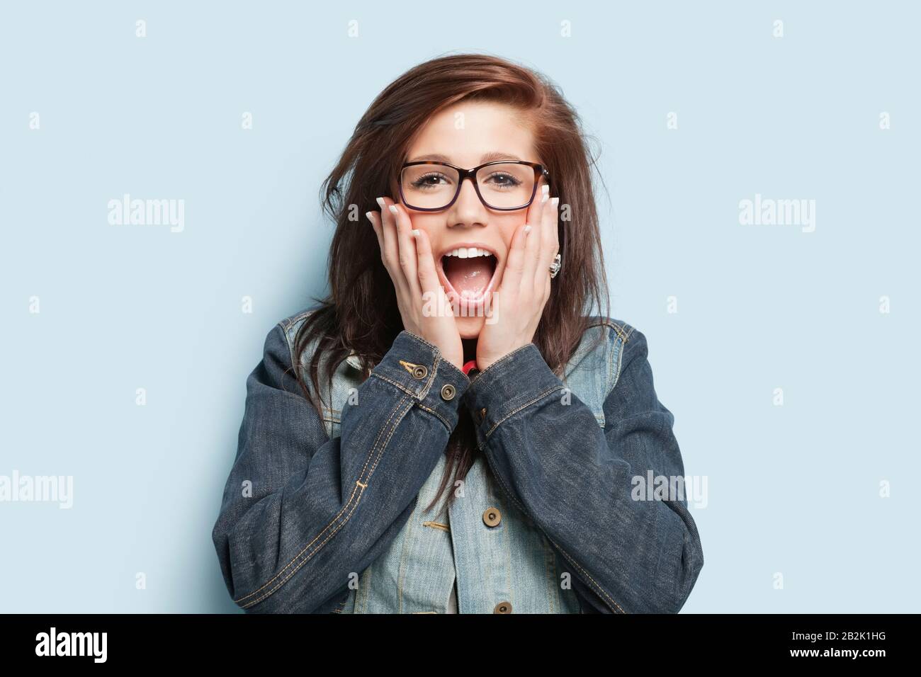 Portrait of surprised young woman with hands on cheeks against light ...