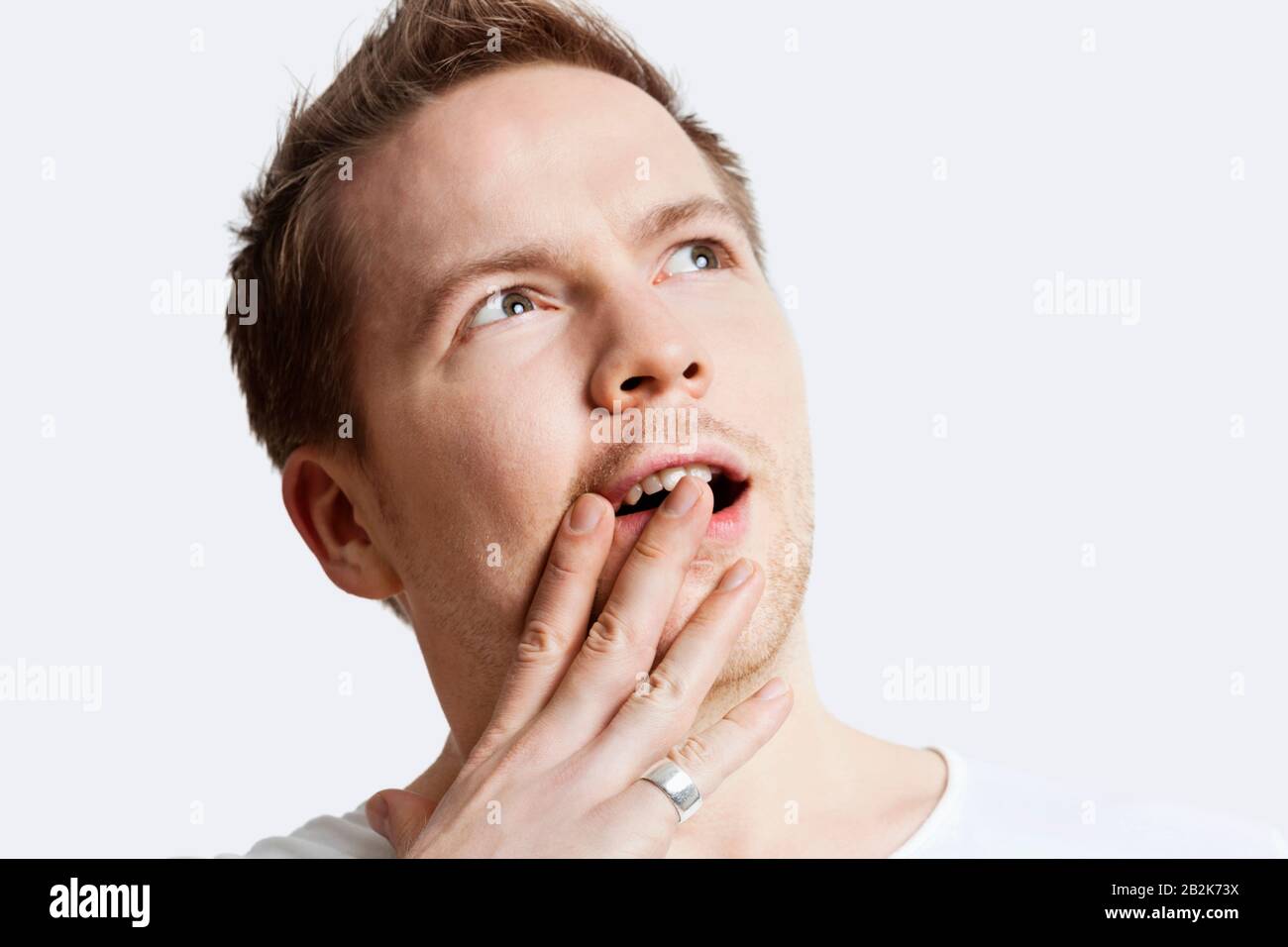 Contemplative young man with hand over mouth against white background ...