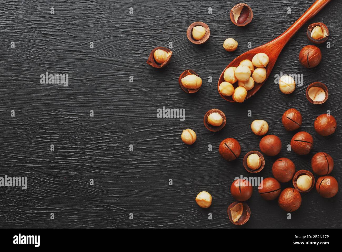 Shelled macadamia nut and peeled macadamia nut on a black textural background in a wooden spoon ...