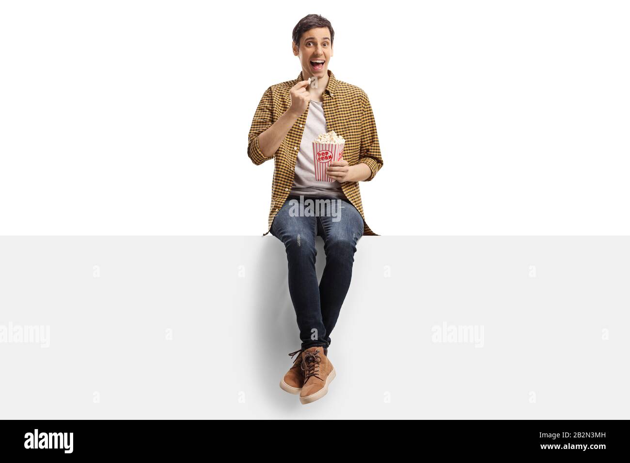Happy young man sitting on a panel and eating popcorn isolated on white ...
