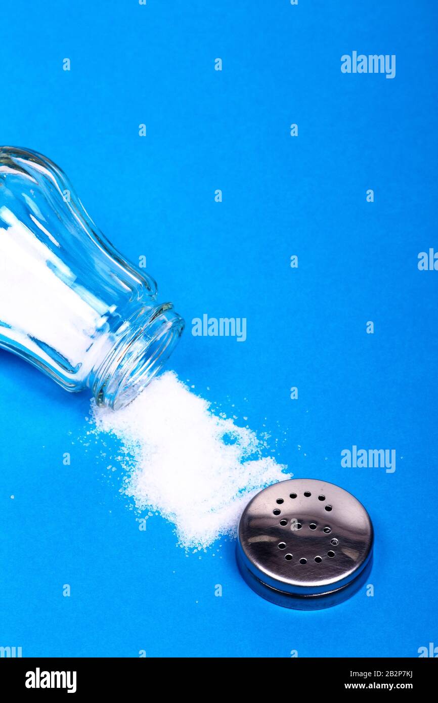 Salt spilling out from a glass sat shaker isolated on a blue background ...