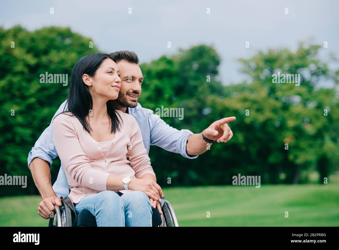 cheerful man pointing with finger while walking in park with disabled ...