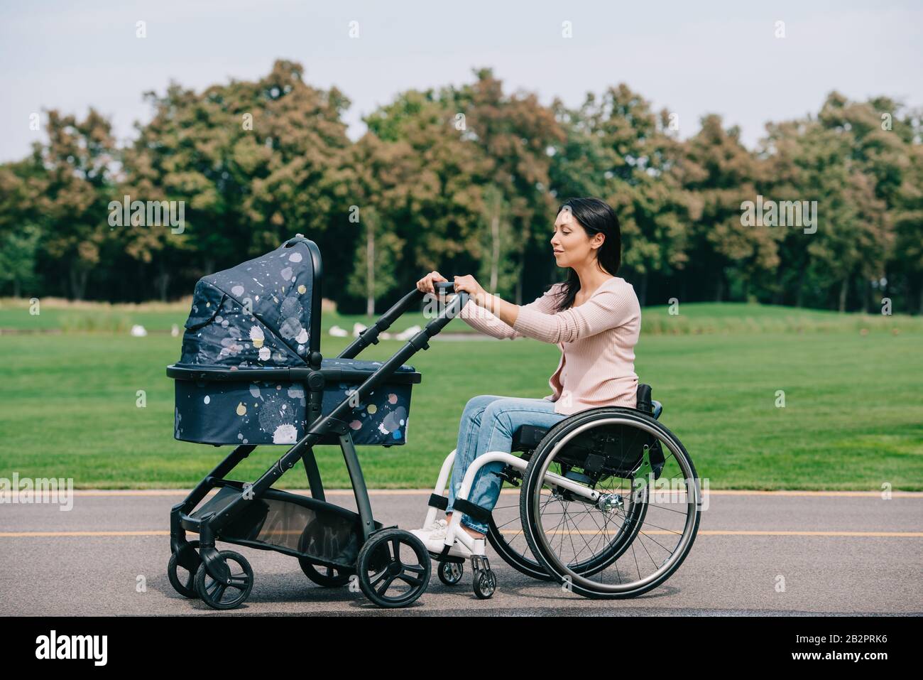 young disabled woman in wheelchair walking with baby carriage in park ...