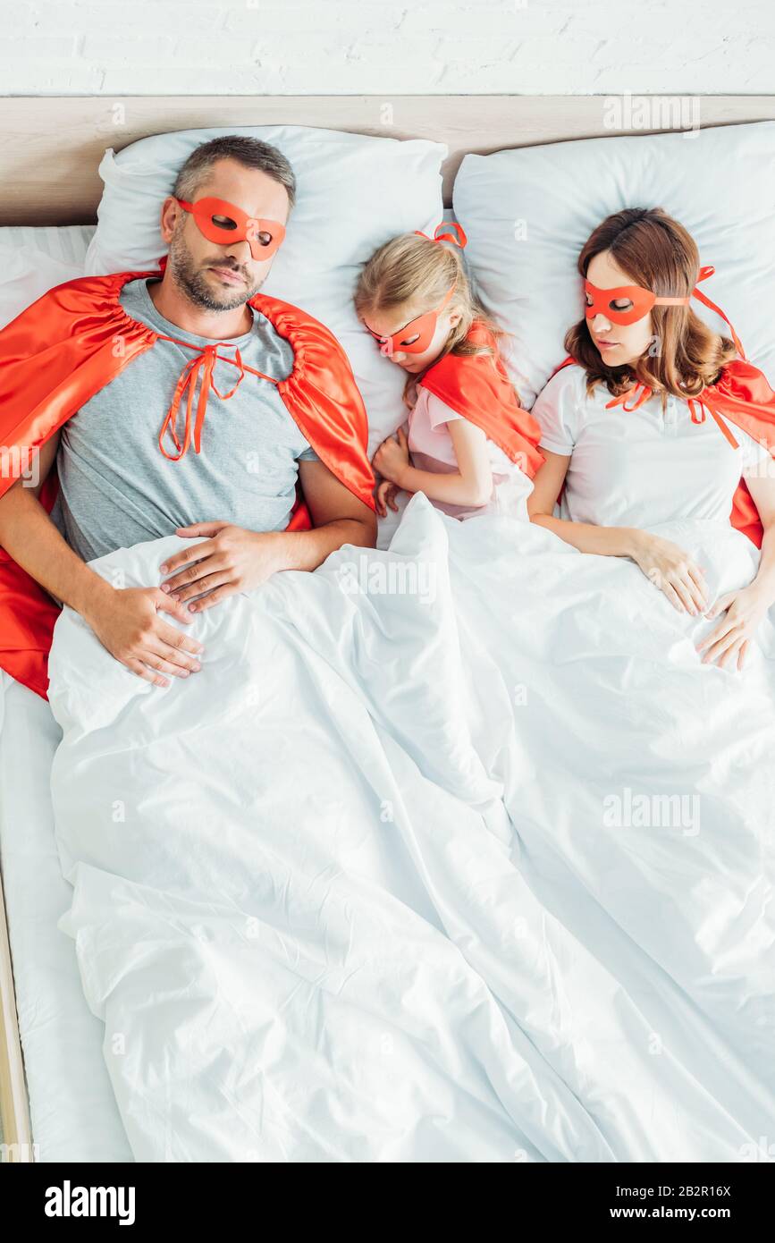 top view of family sleeping on white bedding in costumes of superheroes ...