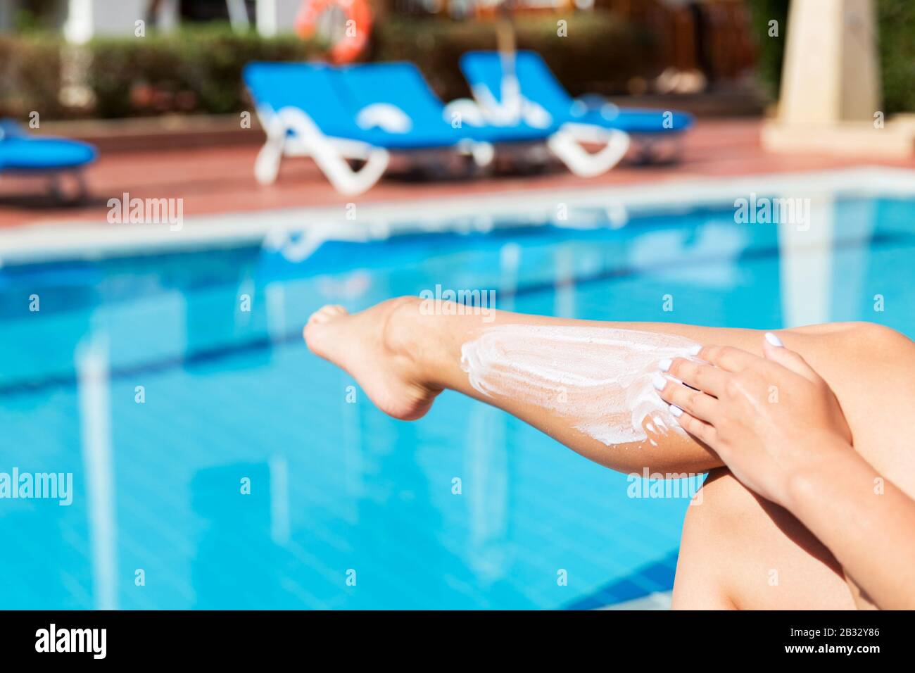 Tanned woman is sitting by the swimming pool and applying sunblock to ...