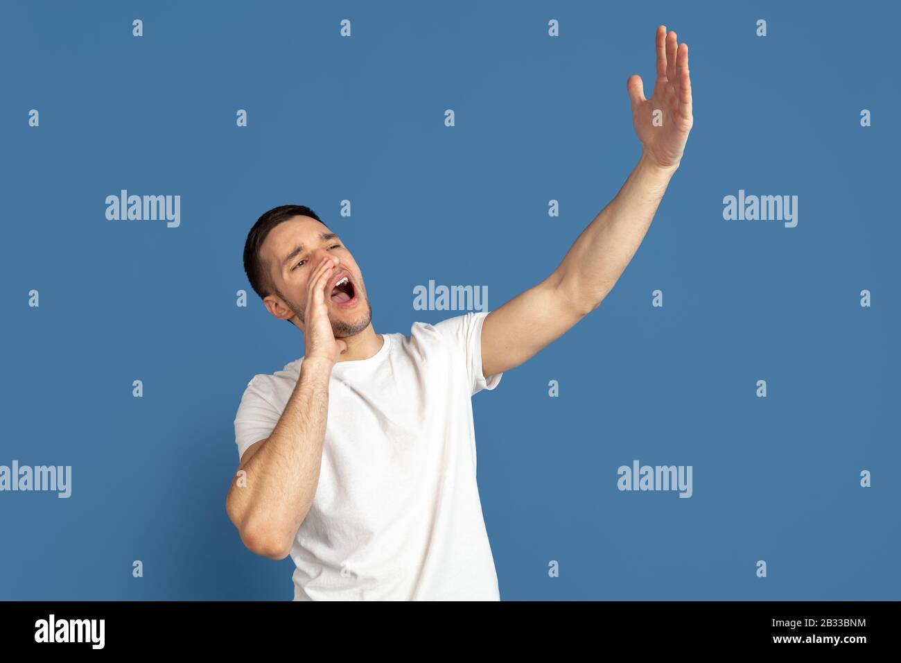 Calling, shouting. Caucasian young man's portrait on blue studio ...
